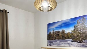 Cama bem arrumada com lençóis brancos e duas almofadas bege, cabeceira de madeira, radiador fixado na parede e um quadro com paisagem nevada.