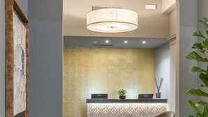 Uma área de recepção com design moderno, com um balcão de atendimento, duas cadeiras, uma mesinha com livros e uma planta grande.