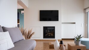 Uma sala de estar moderna com um sofá cinza, uma mesa de centro de madeira, uma televisão de tela plana na parede e uma lareira abaixo da TV.