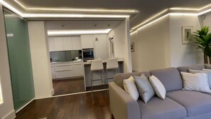 Uma sala de estar moderna com um sofá cinzento e uma cozinha ao fundo.