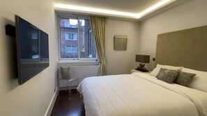 Quarto de hotel com cama, televisão na parede, janela com cortinas, mesa de cabeceira com abajur e vista para edifícios.