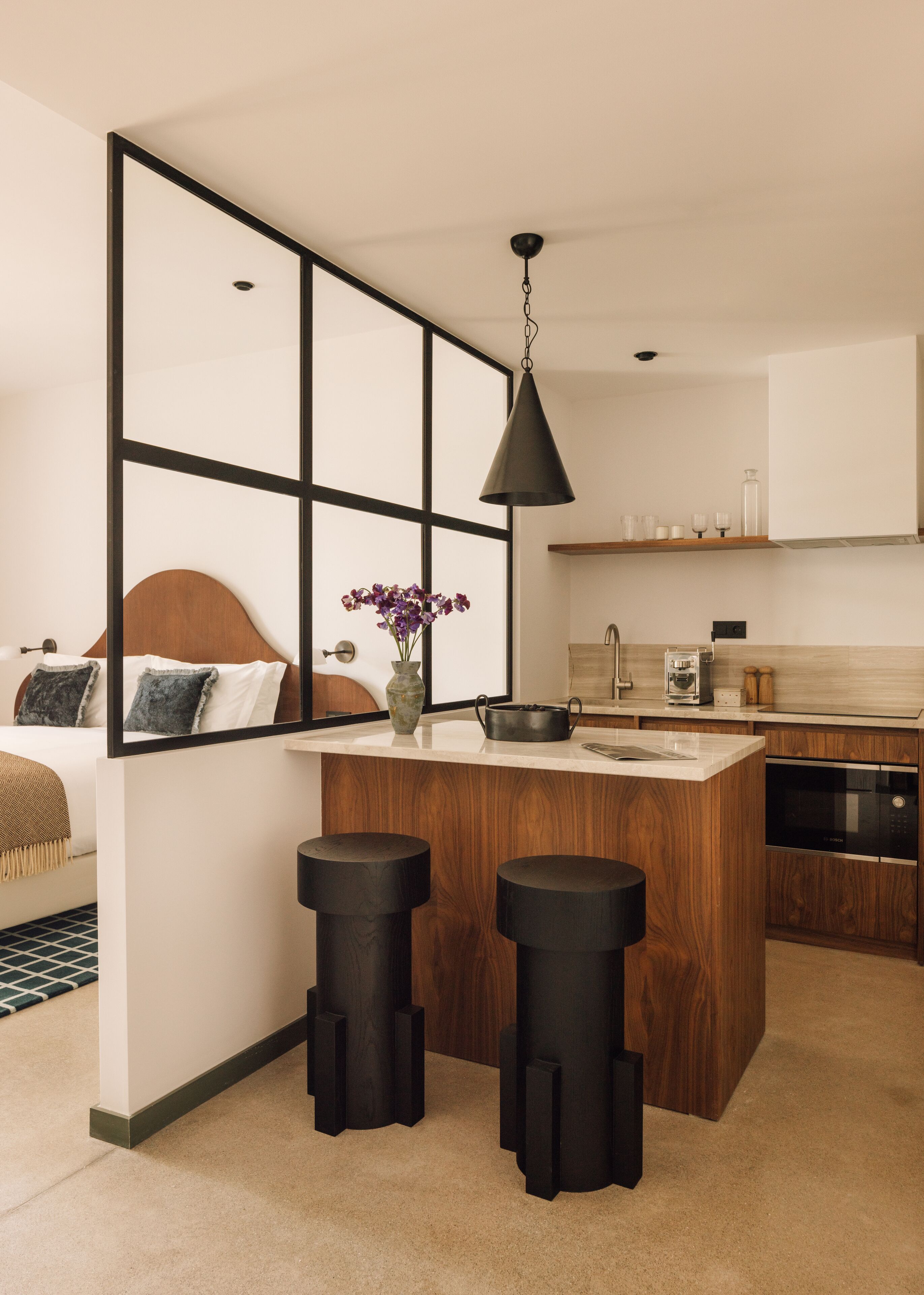 Cozinha moderna com ilha central, banquetas pretas, luminária pendente e cabeceira de madeira no quarto vizinho.