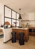 Cozinha moderna com ilha central, banquetas pretas, luminária pendente e cabeceira de madeira no quarto vizinho.