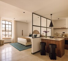 Quarto de hotel moderno com uma cama grande, um espaço de estar pequeno, uma kitchenette e uma janela ampla com cortinas.