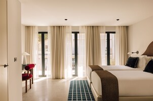 Quarto de hotel com duas camas, uma cadeira vermelha, uma luminária e vista para o exterior por grandes janelas.