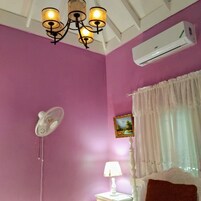 Quarto com paredes roxas, ar condicionado branco, ventilador de teto, luminária de cabeceira e uma cama com cortinas brancas.