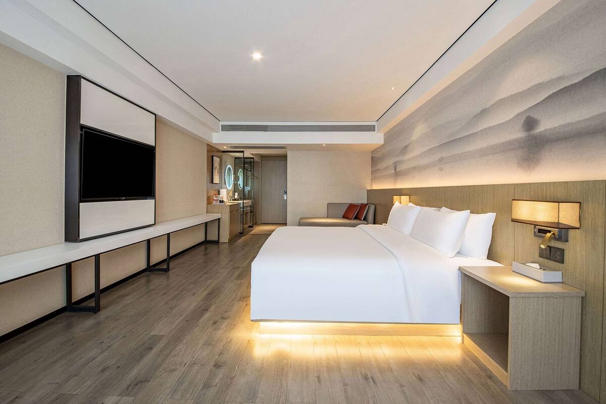 Quarto de hotel moderno com uma cama grande, televisão de tela plana, um banco, uma mesa de trabalho e um banheiro ao fundo.
