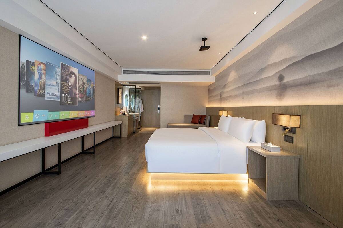 Quarto de hotel moderno com uma grande televisão de tela plana exibindo pôsteres de filmes, uma cama bem arrumada, uma mesa de cabeceira com abajur e uma cabeceira de madeira.