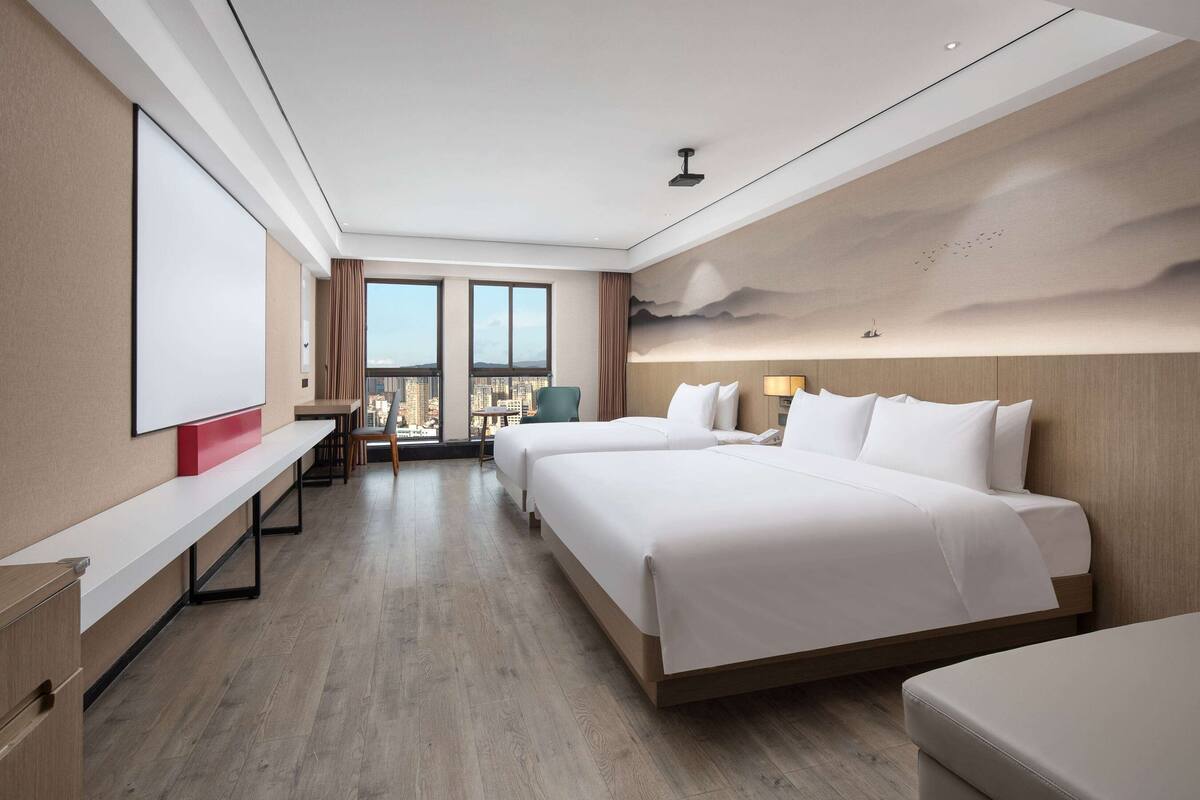 Quarto de hotel com uma cama grande, uma escrivaninha e vista da cidade através de amplas janelas.