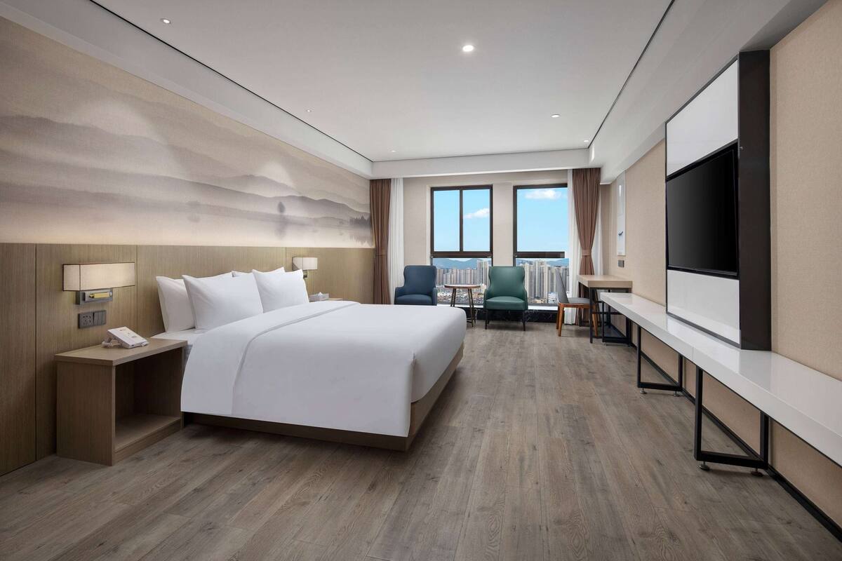 Quarto de hotel com uma cama grande, uma escrivaninha, duas cadeiras, televisão e vista para a cidade.