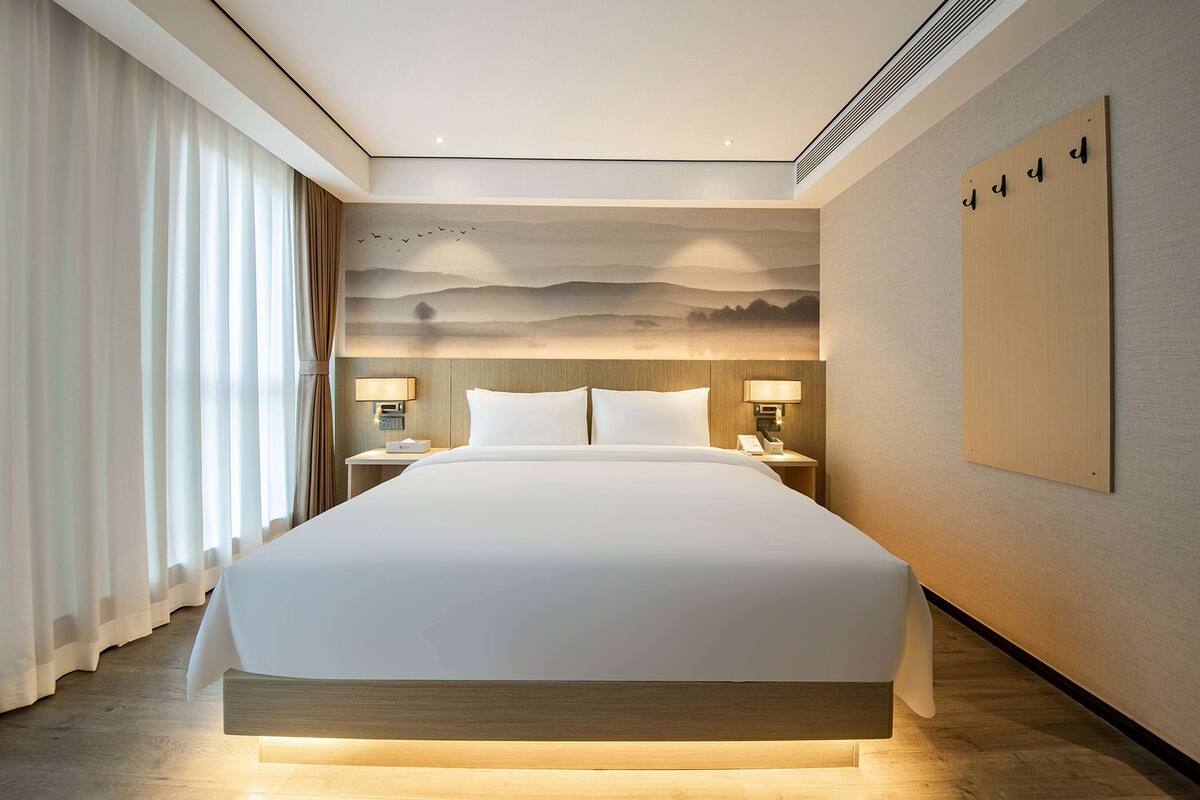 Quarto de hotel moderno com uma cama grande, luminárias laterais, cabeceira de madeira e cabideiro fixado na parede.