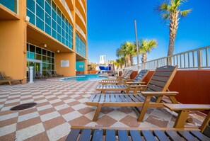 Uma área à beira da piscina com espreguiçadeiras e piso xadrez, com vista para um edifício com amplas janelas e palmeiras.