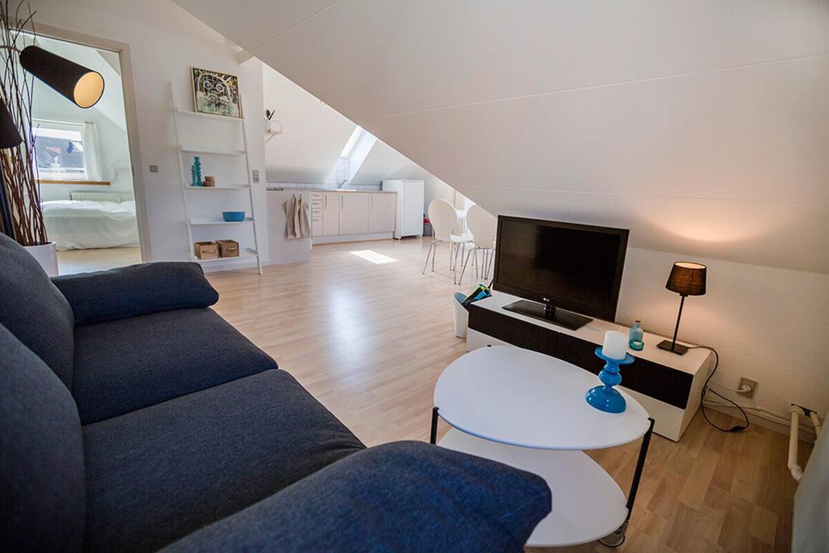 Um apartamento moderno com sala de estar, que inclui sofá, TV e uma pequena mesa. Há uma cozinha com armários brancos e uma mesa de jantar. O apartamento possui teto inclinado e grandes janelas.