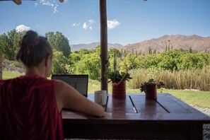 Uma pessoa sentada a uma mesa de madeira com um laptop, uma caneca e duas plantas em vasos. A mesa está ao ar livre, com vista para árvores verdes e montanhas ao fundo.
