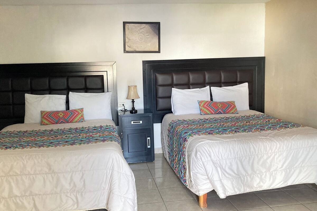 Quarto de hotel com duas camas, cada uma com cabeceira, roupa de cama branca e colcha estampada. Há uma mesa de cabeceira com abajur e um pequeno armário entre as camas.