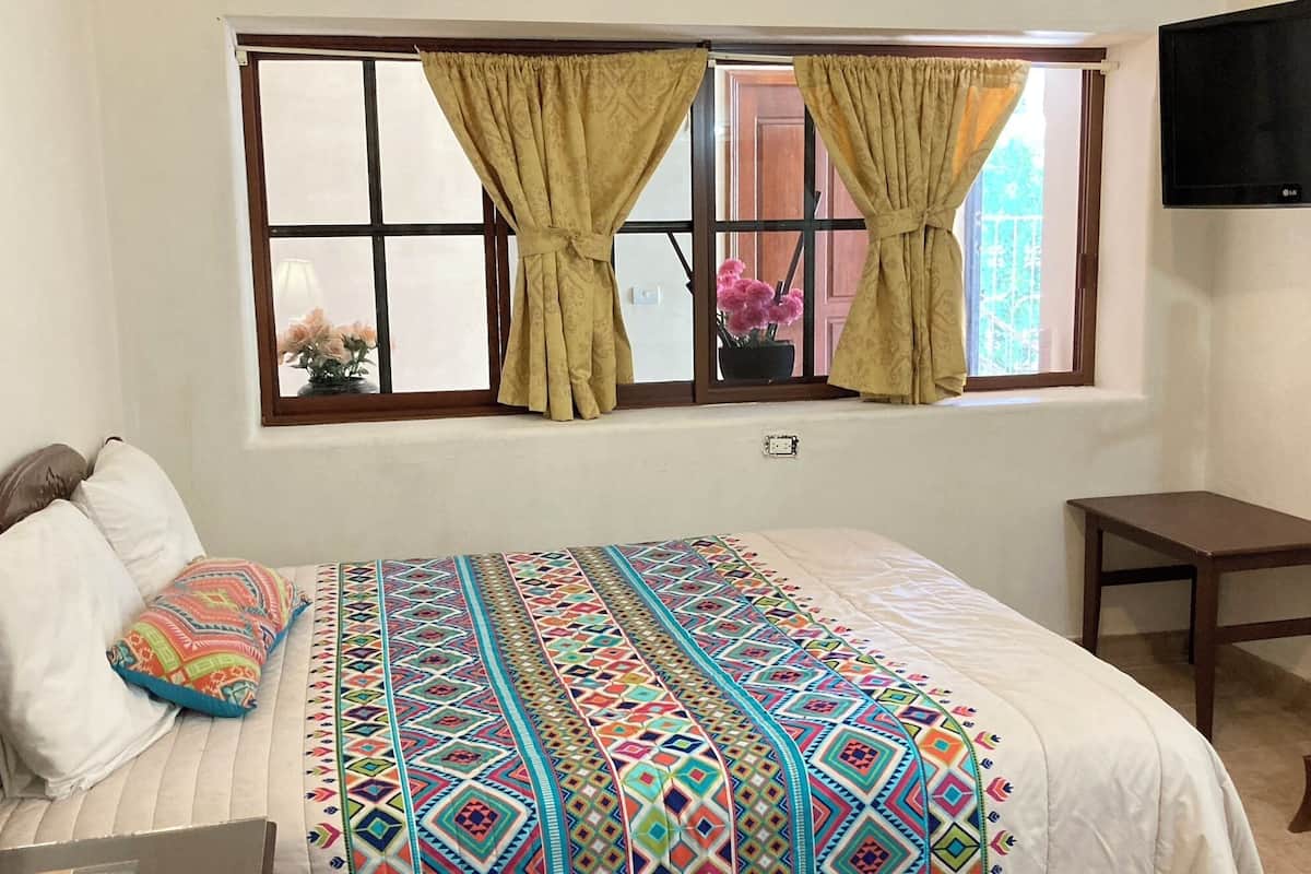 Quarto com cama, almofadas, janela com cortinas, televisão e uma mesinha.