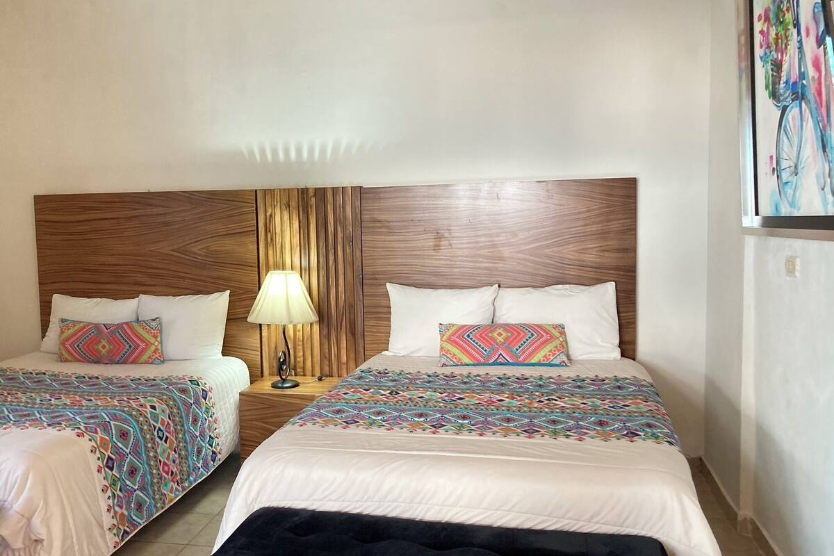 Quarto de hotel com duas camas, cada uma com colcha e almofadas estampadas, cabeceira de madeira e mesa de cabeceira com luminária.