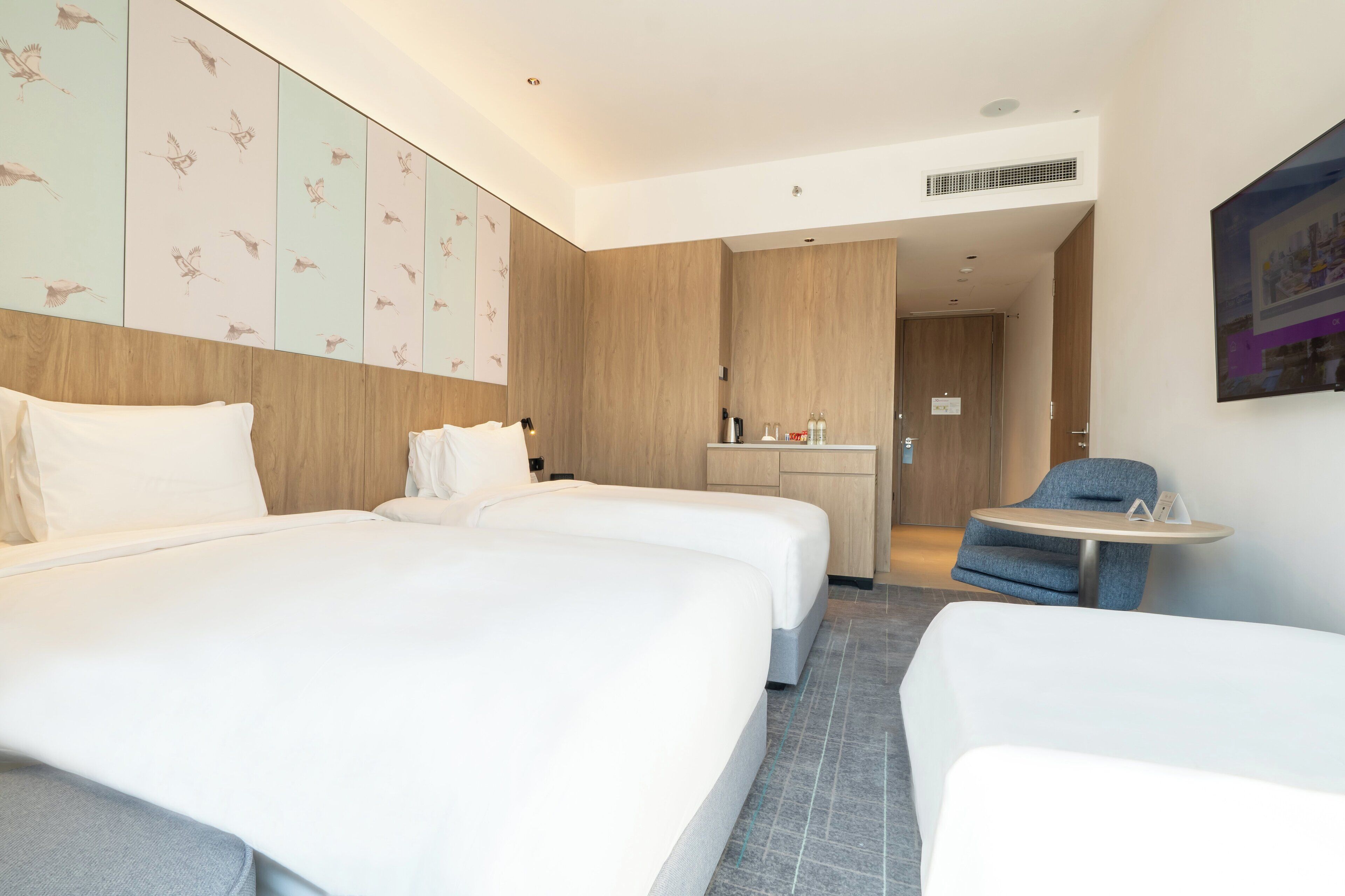 Quarto de hotel com duas camas, uma escrivaninha e uma cadeira. Há uma mesinha com um telefone e uma chávena. O quarto tem painéis de madeira e uma parede com um padrão de pássaros.