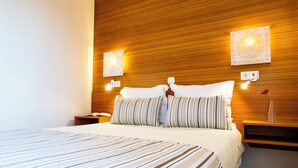 Quarto de hotel com cabeceira de madeira, cama grande com roupa de cama listrada, duas mesas de cabeceira com abajures e luminária fixada na parede.