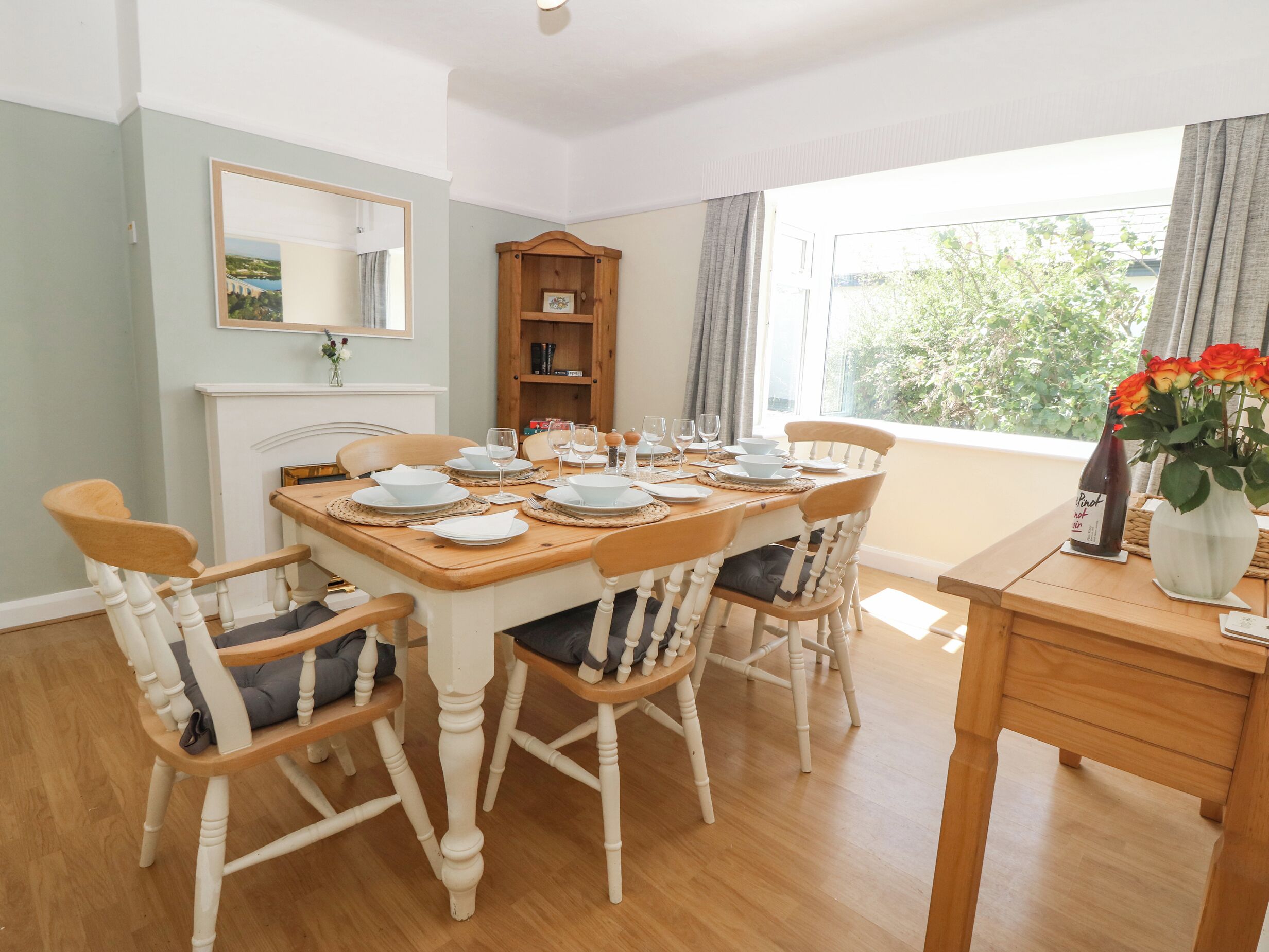 Uma sala de jantar com uma mesa de madeira preparada para seis pessoas, rodeada por cadeiras brancas com almofadas escuras. Um armário de madeira com um espelho acima da mesa e uma janela com cortinas à direita.