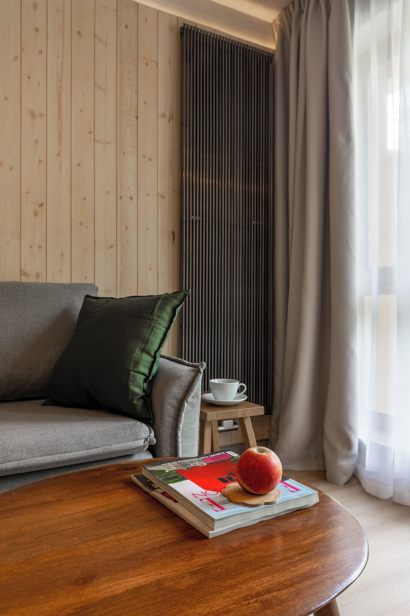 Um quarto acolhedor com parede de madeira, um sofá cinzento, uma mesa de centro de madeira com um livro e uma maçã, e uma pequena mesa branca com uma chávena.