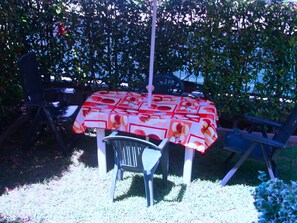 Uma mesa com toalha florida, rodeada por cadeiras, em ambiente externo com árvores ao fundo.