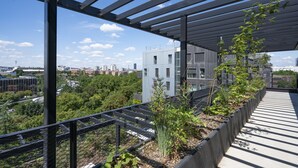 Um jardim no terraço com uma pérgola de metal, cercado por vegetação e edifícios da cidade ao fundo.