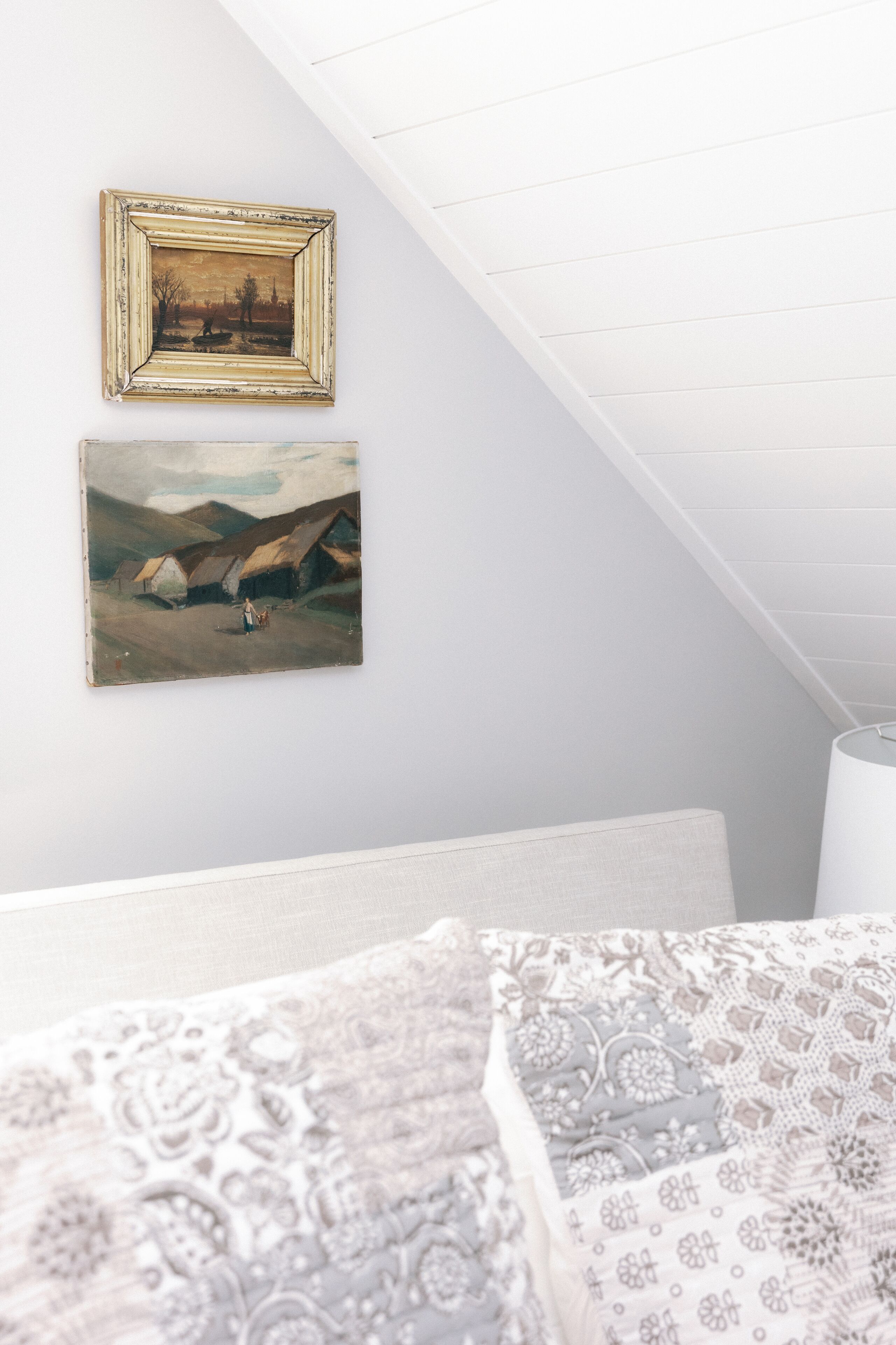 Quarto com teto inclinado, duas pinturas na parede e roupa de cama estampada.