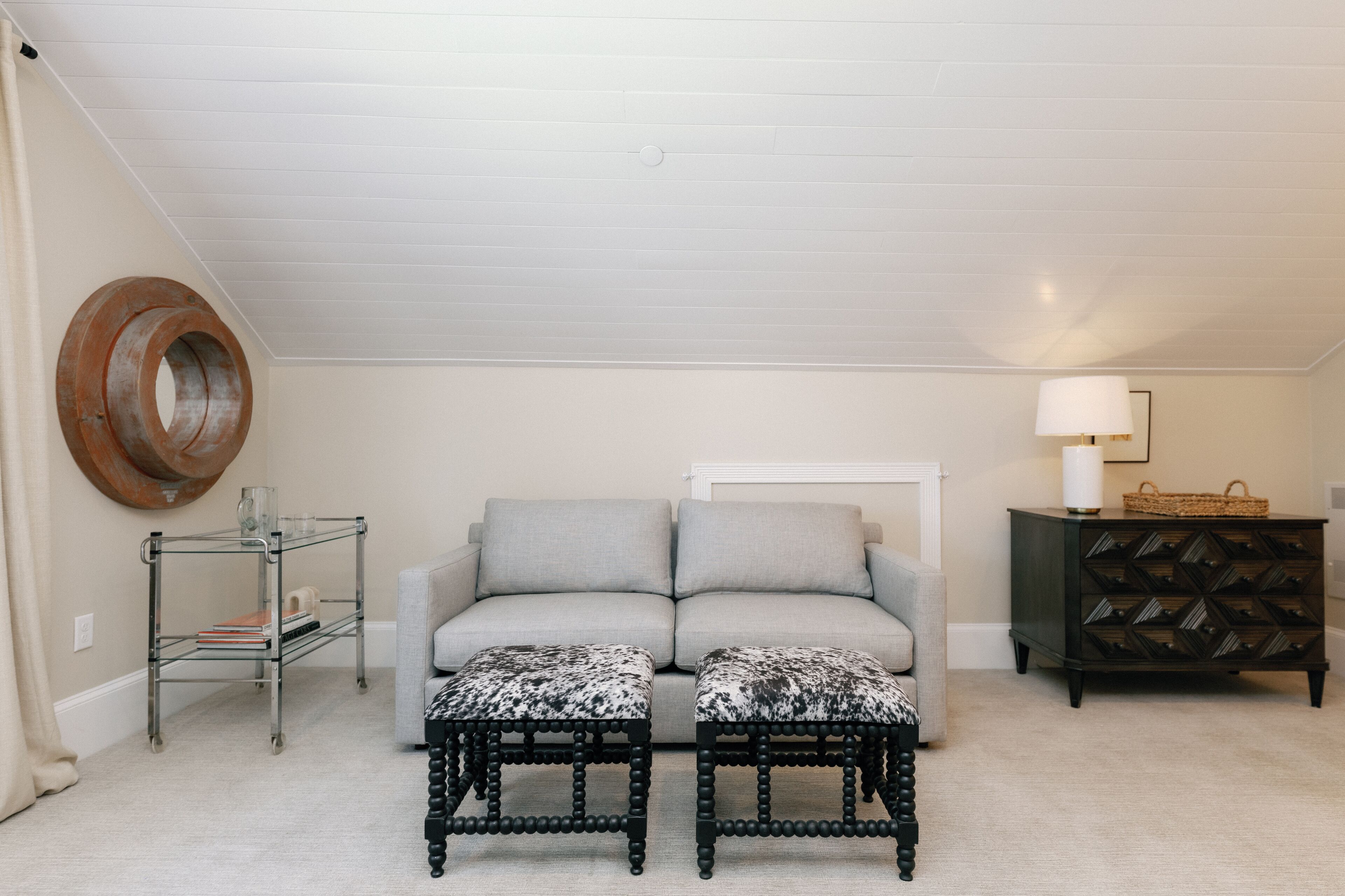 Uma sala de estar com um sofá cinzento, dois banquinhos estofados preto e branco, uma mesinha lateral de metal com livros, outra mesinha de madeira com um abajur e uma decoração circular na parede.