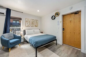 Quarto com uma cama, uma poltrona azul, uma porta de madeira e uma janela com persianas.