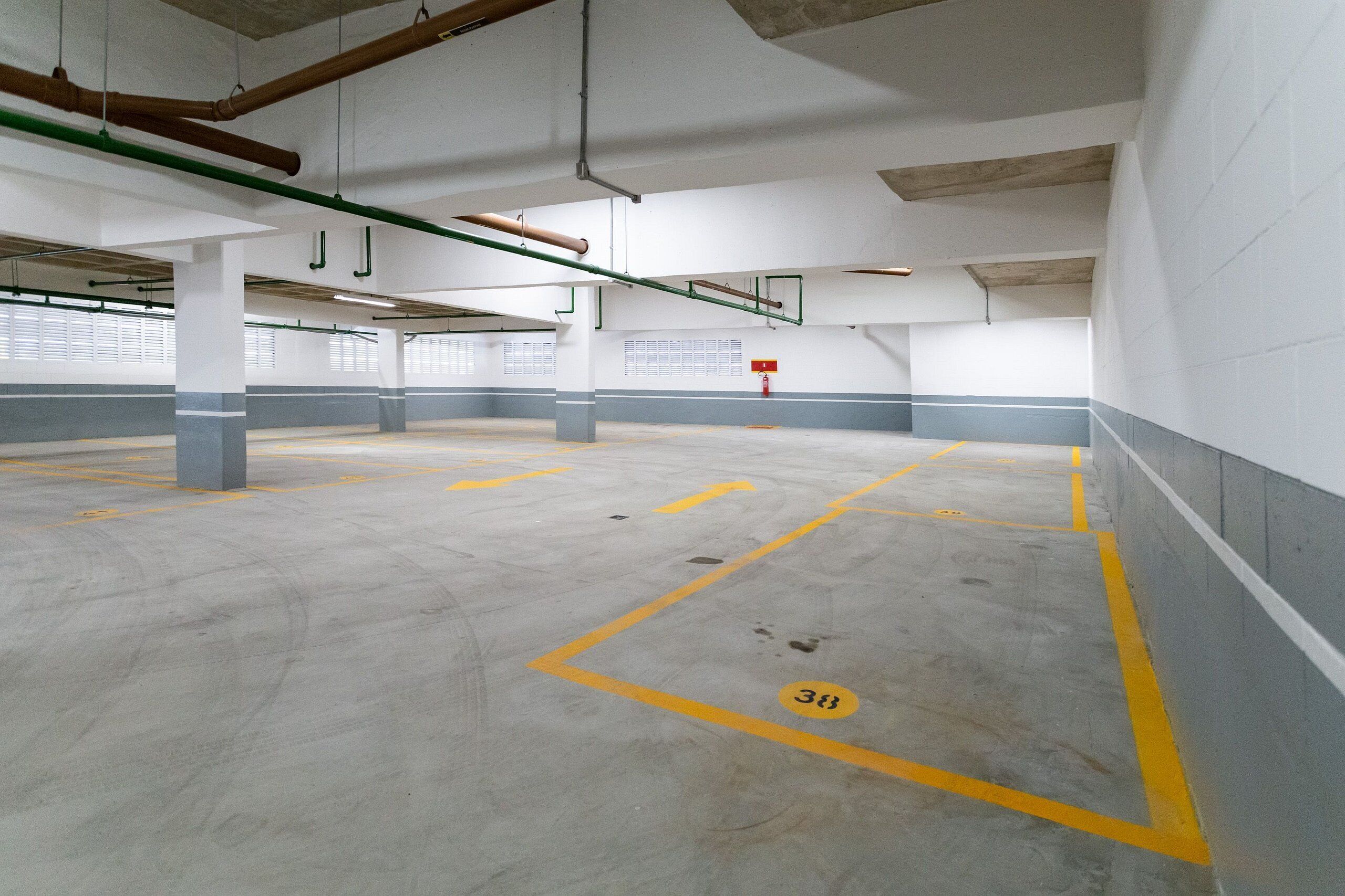 Garagem de estacionamento vazia, com pisos de concreto, linhas de estacionamento amarelas e tubulações verdes no teto.