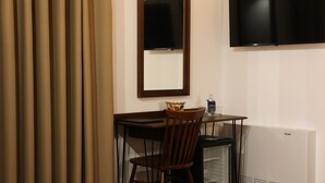 Quarto compacto com escrivaninha de madeira, uma cadeira, frigorífico pequeno e televisão fixada na parede.
