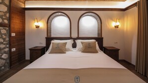 Quarto de hotel com uma cama grande, duas mesas de cabeceira, parede de pedra, teto de madeira e janelas em arco.