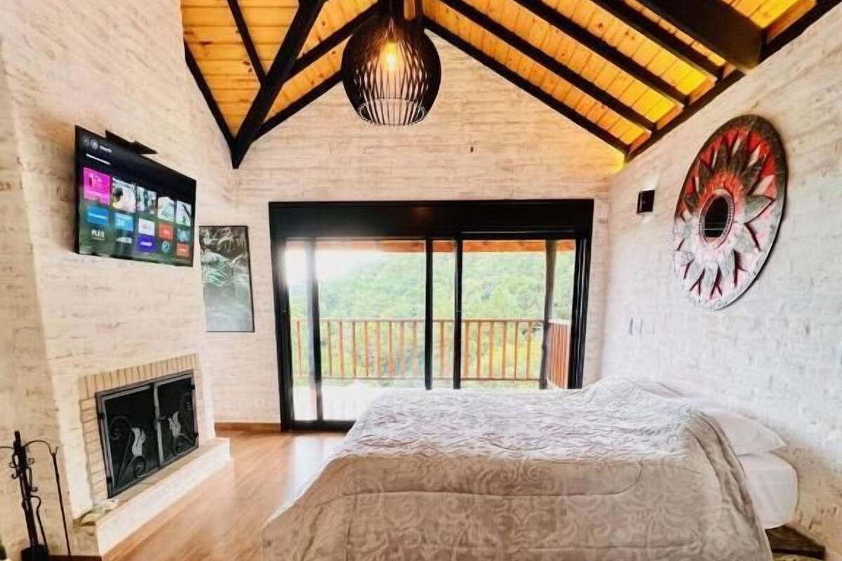 Quarto com cama grande, televisão na parede, lareira e varanda com vista para o verde.