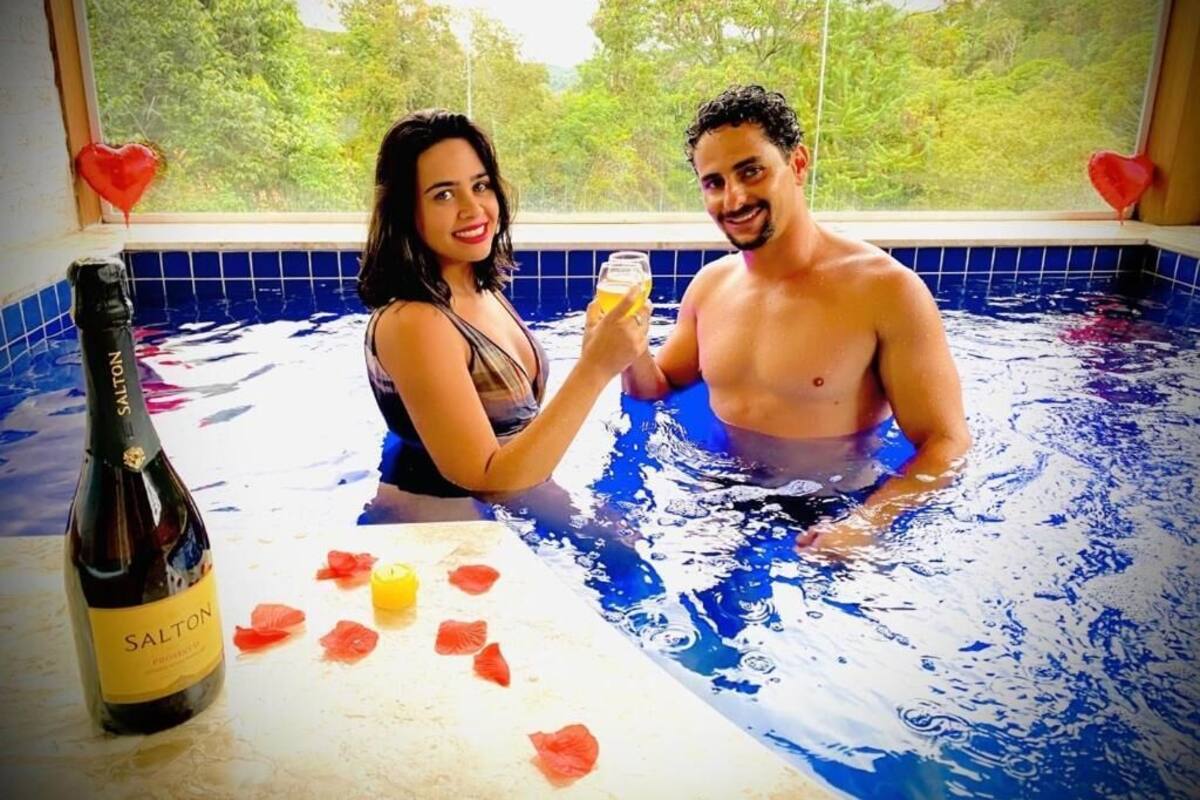 Um casal em uma banheira de hidromassagem com uma garrafa de vinho Salton e pétalas de rosa na borda.