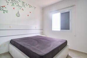 Quarto com cama, janela com persianas, interruptor de luz na parede e um adorno de gaiola de pássaro com folhas.