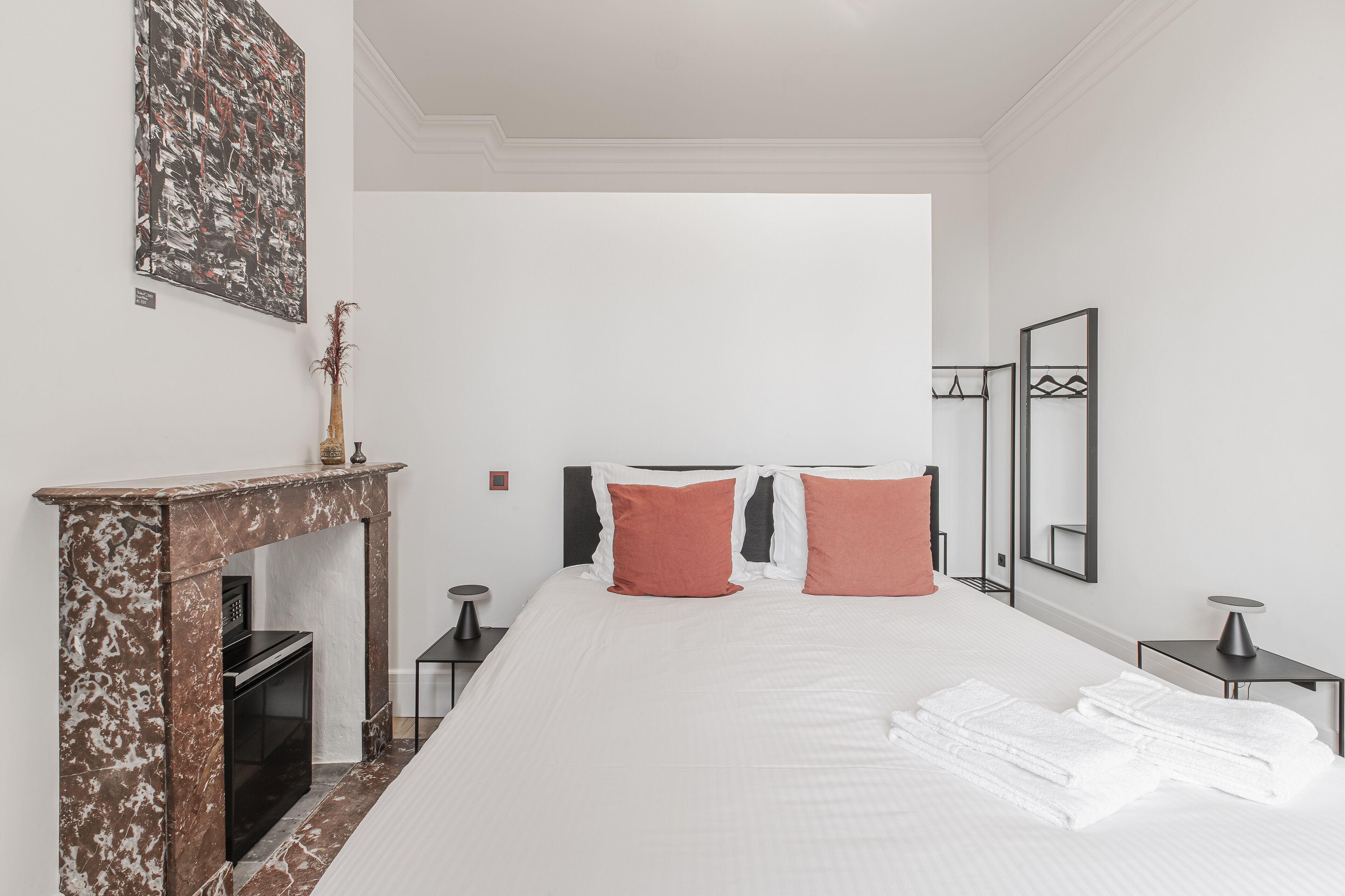 Quarto de hotel com uma cama grande, duas almofadas vermelhas, cabeceira branca, lareira de mármore, uma mesa pequena e um espelho na parede.
