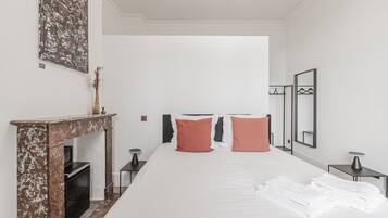 Quarto de hotel com uma cama grande, duas almofadas vermelhas, cabeceira branca, lareira de mármore, uma mesa pequena e um espelho na parede.