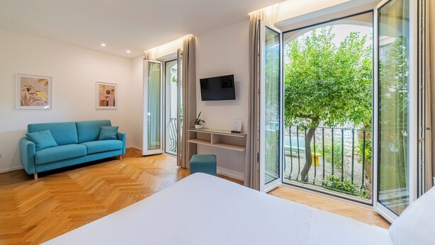 Quarto moderno com uma cama grande, um sofá azul, uma televisão e uma porta de vidro deslizante para uma varanda com uma árvore.