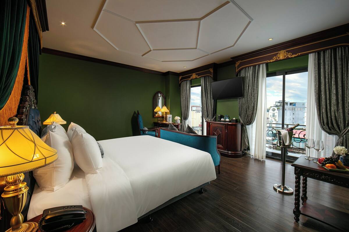 Quarto de hotel com uma cama grande, uma mesa com abajur, uma televisão e uma varanda com vista para a cidade.