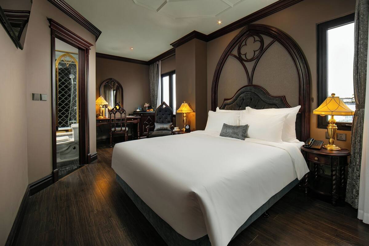 Quarto de hotel espaçoso com uma cama grande, duas mesas de cabeceira com abajures, um espelho e vista da cidade pela janela.