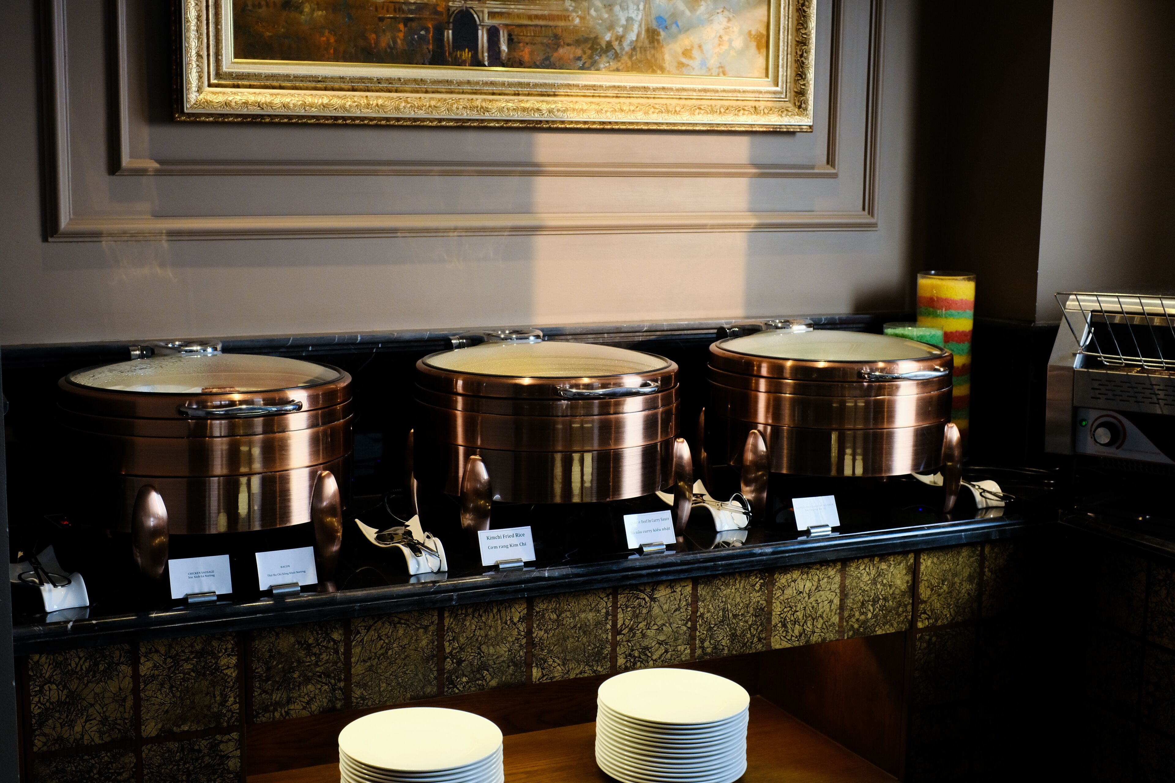 Uma área de buffet com três grandes recipientes de cobre para buffet, identificados com diferentes tipos de arroz, uma pilha de pratos brancos e uma vela colorida.
