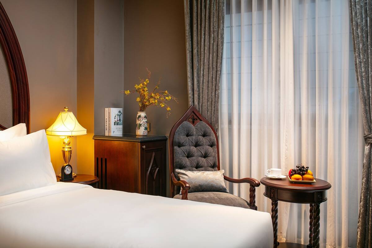 Quarto de hotel com cama, luminária, cadeira, mesinha de centro com uma fruteira e um vaso com flores.