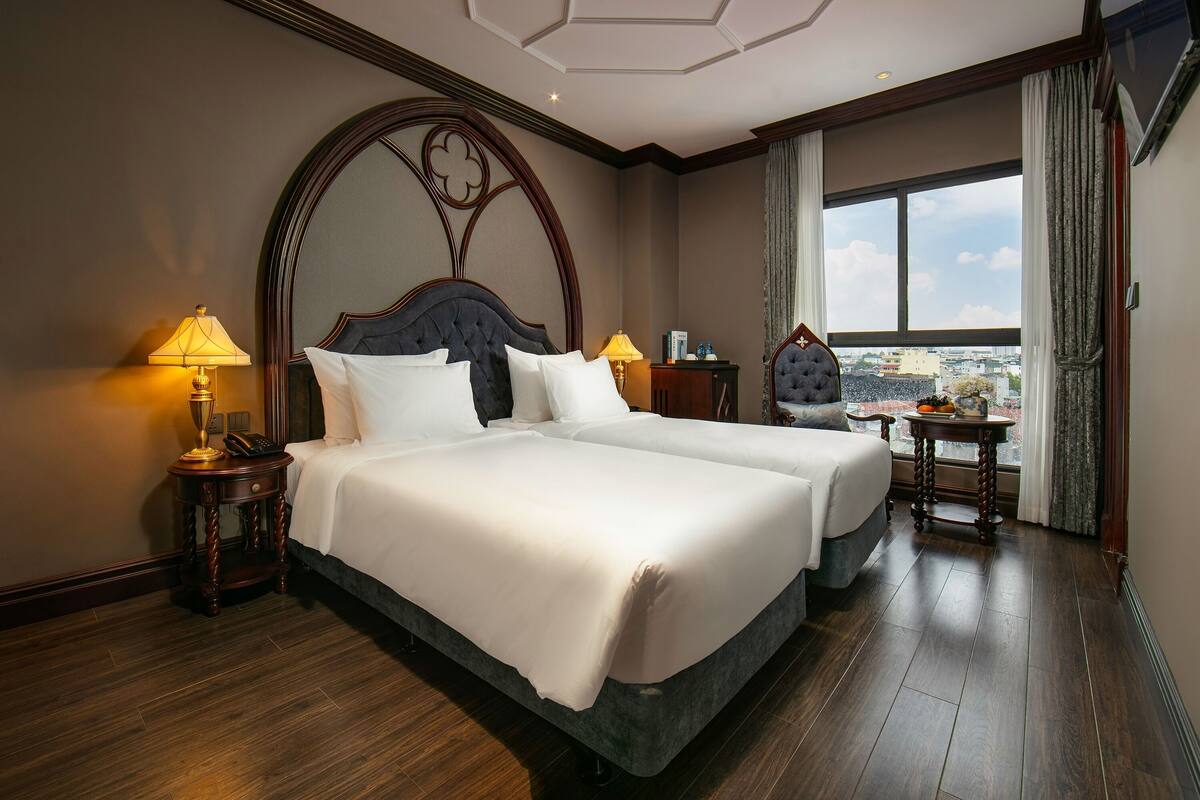 Quarto de hotel com duas camas, uma janela grande com vista para a cidade, um espelho redondo acima da cabeceira e uma pequena mesa com uma luminária.
