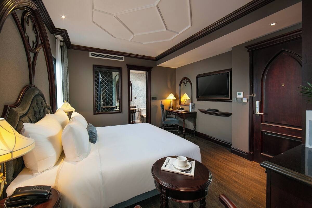 Quarto de hotel com uma cama grande, abajures, escrivaninha com cadeira, televisão e guarda-roupa.