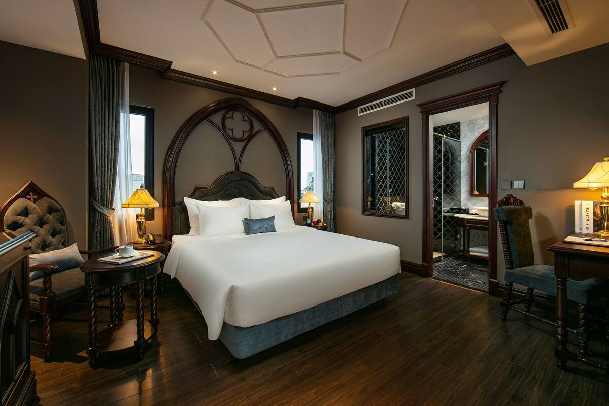 Um quarto de hotel espaçoso com uma cama grande, cabeceira de madeira, área de estar com uma cadeira e mesa, e vista através de uma porta em arco.