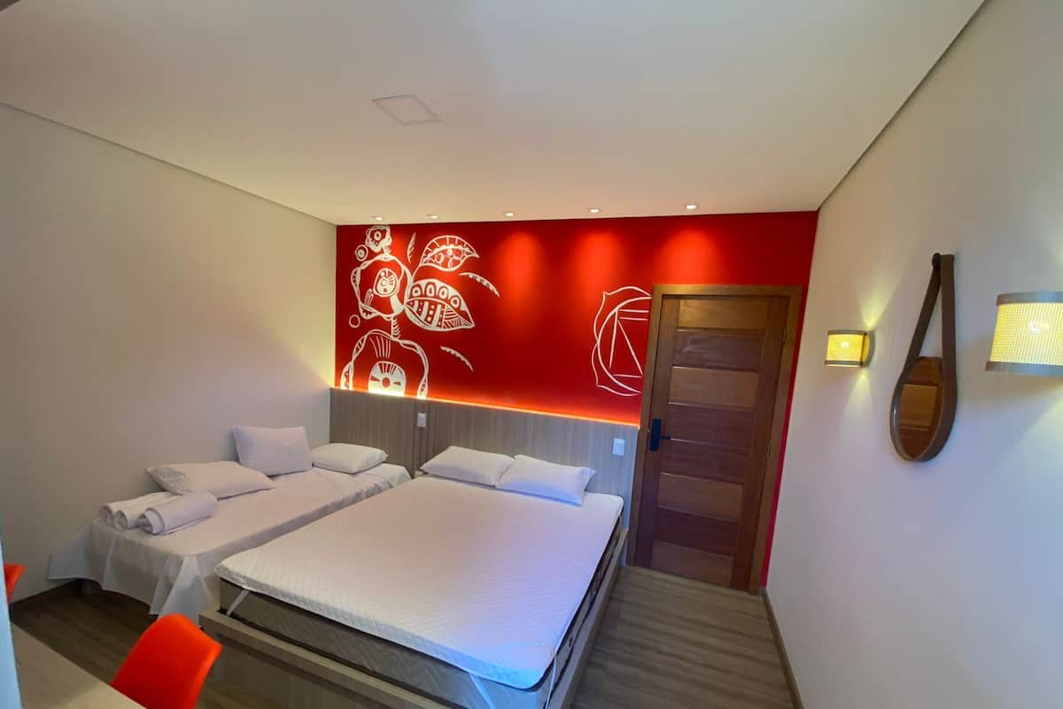 Quarto de hotel moderno com duas camas, piso de madeira e parede em vermelho com padrões brancos.