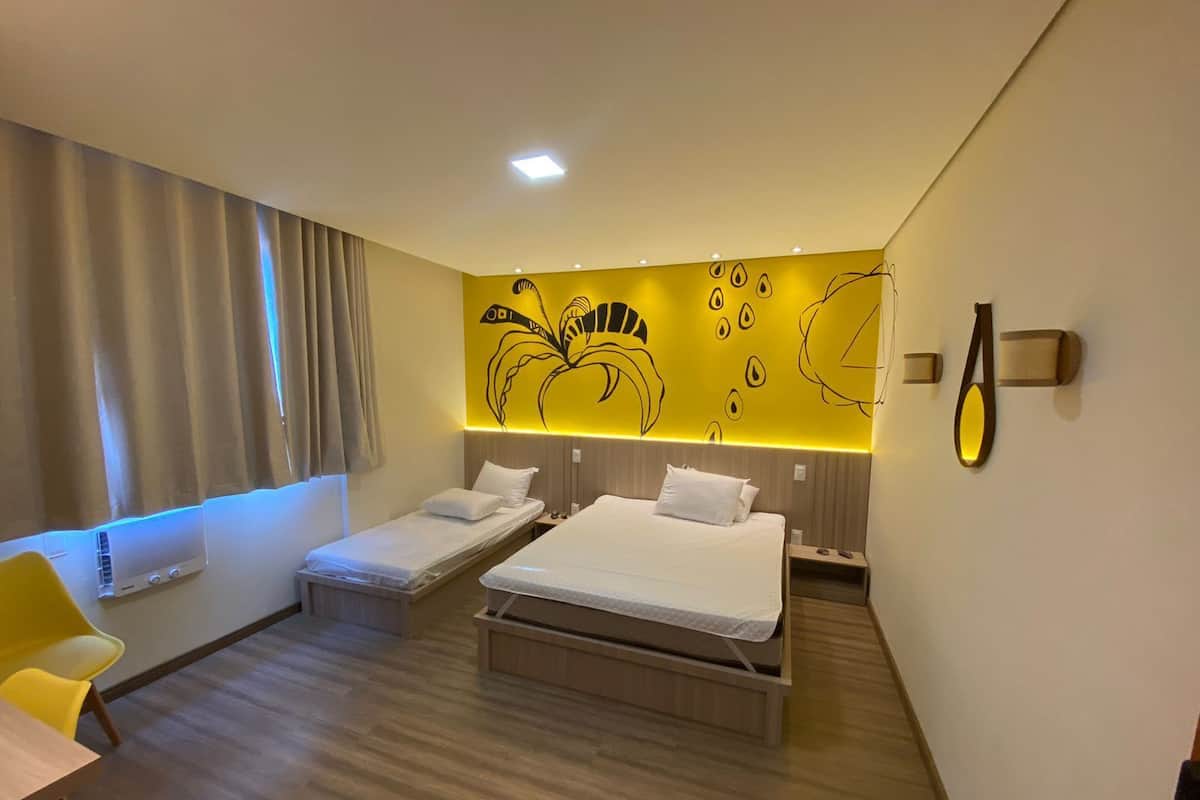 Quarto de hotel com duas camas de solteiro, uma mesinha, uma cadeira, uma luminária na parede e uma janela com cortinas.