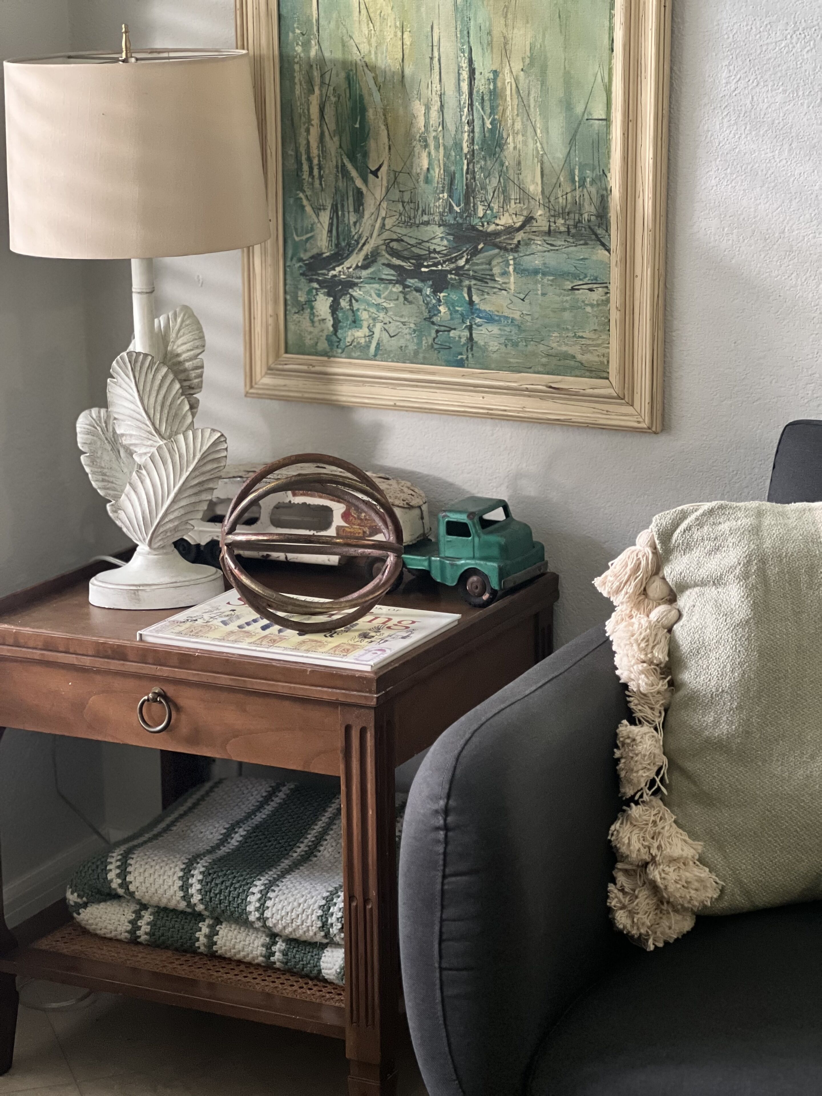 Uma mesa lateral de madeira com uma luminária, um rádio antigo e um quadro emoldurado com uma cena de floresta.