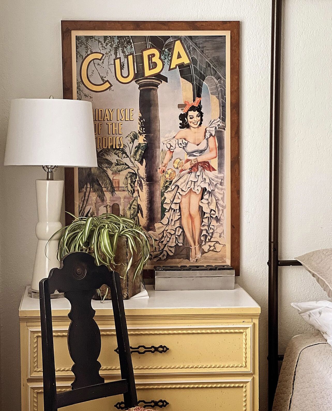 Um cartaz antigo emoldurado retrata uma mulher em vestido, com a palavra 'CUBA' em uma cômoda amarela com gavetas, uma luminária branca e uma cadeira preta.